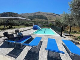 Piscina com montanhas ao fundo, rodeada por espreguiçadeiras e um espaço de estar coberto.