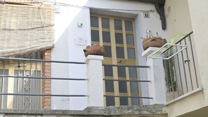 Uma varanda com guarda-corpo de metal, uma janela com porta de madeira e duas plantas em vasos no corrimão.