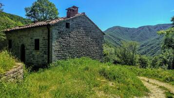 Um edifício de pedra com telhado em tijolos, rodeado por vegetação exuberante e montanhas ao fundo.