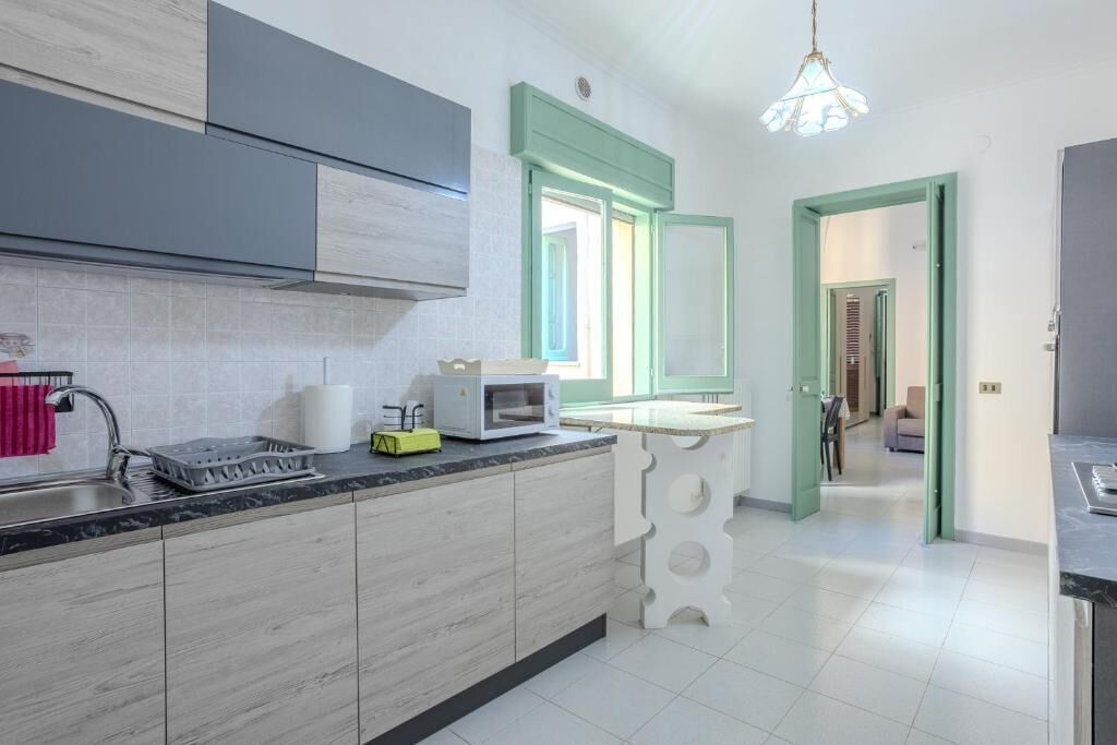 Uma cozinha moderna com armários cinza, bancada branca e micro-ondas. Há uma janela com moldura verde e um lustre.