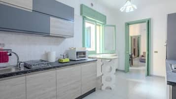 Uma cozinha moderna com armários cinza, bancada branca e micro-ondas. Há uma janela com moldura verde e um lustre.