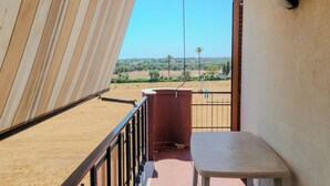 Uma varanda com vista para uma paisagem rural, uma mesa pequena e uma lixeira vermelha.