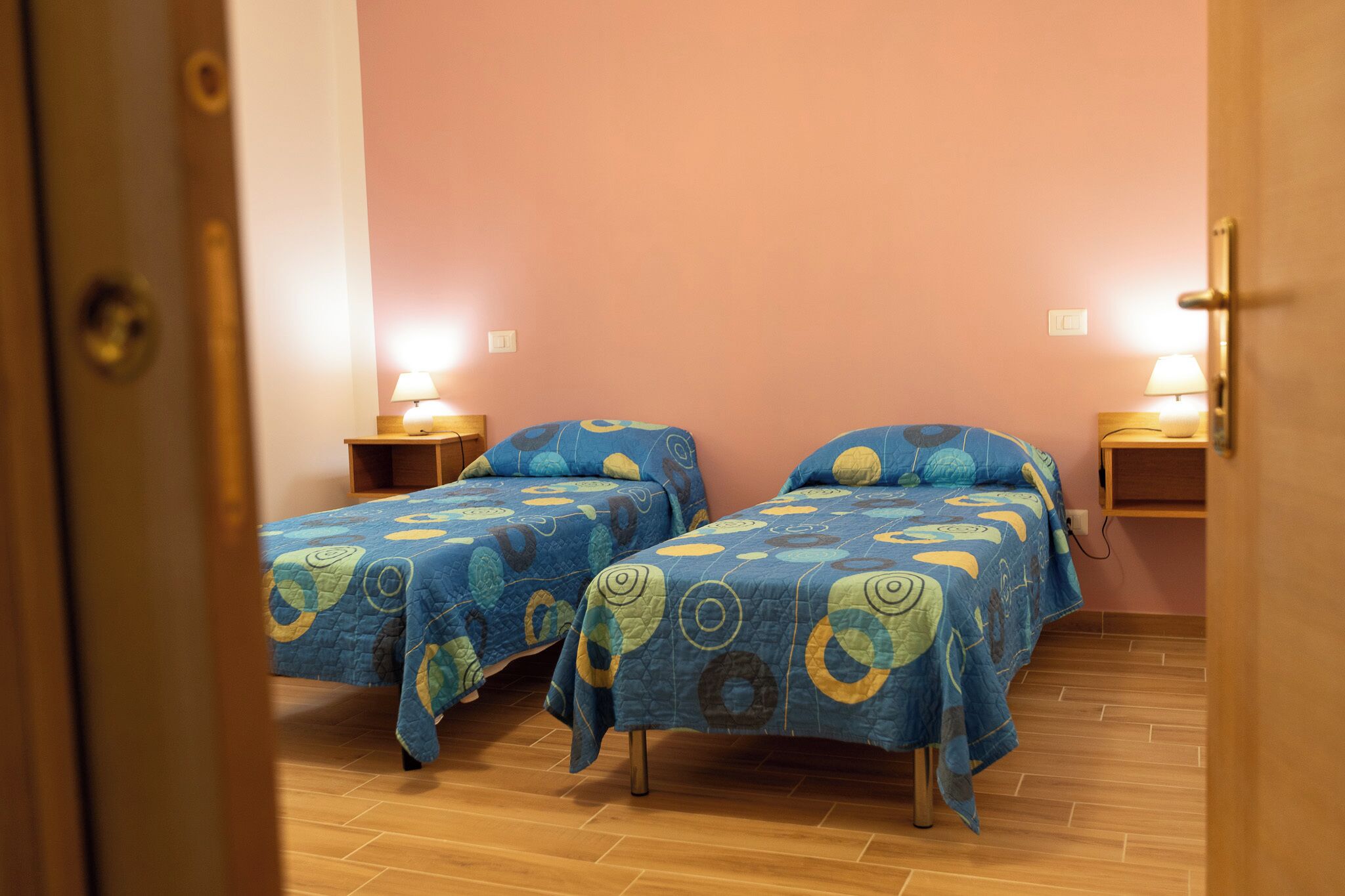 Quarto com duas camas de solteiro, cada uma com colcha estampada azul e almofada combinando. Há mesas de cabeceira com luminárias e o piso é de madeira.