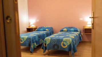 Quarto com duas camas de solteiro, cada uma com colcha estampada azul e almofada combinando. Há mesas de cabeceira com luminárias e o piso é de madeira.