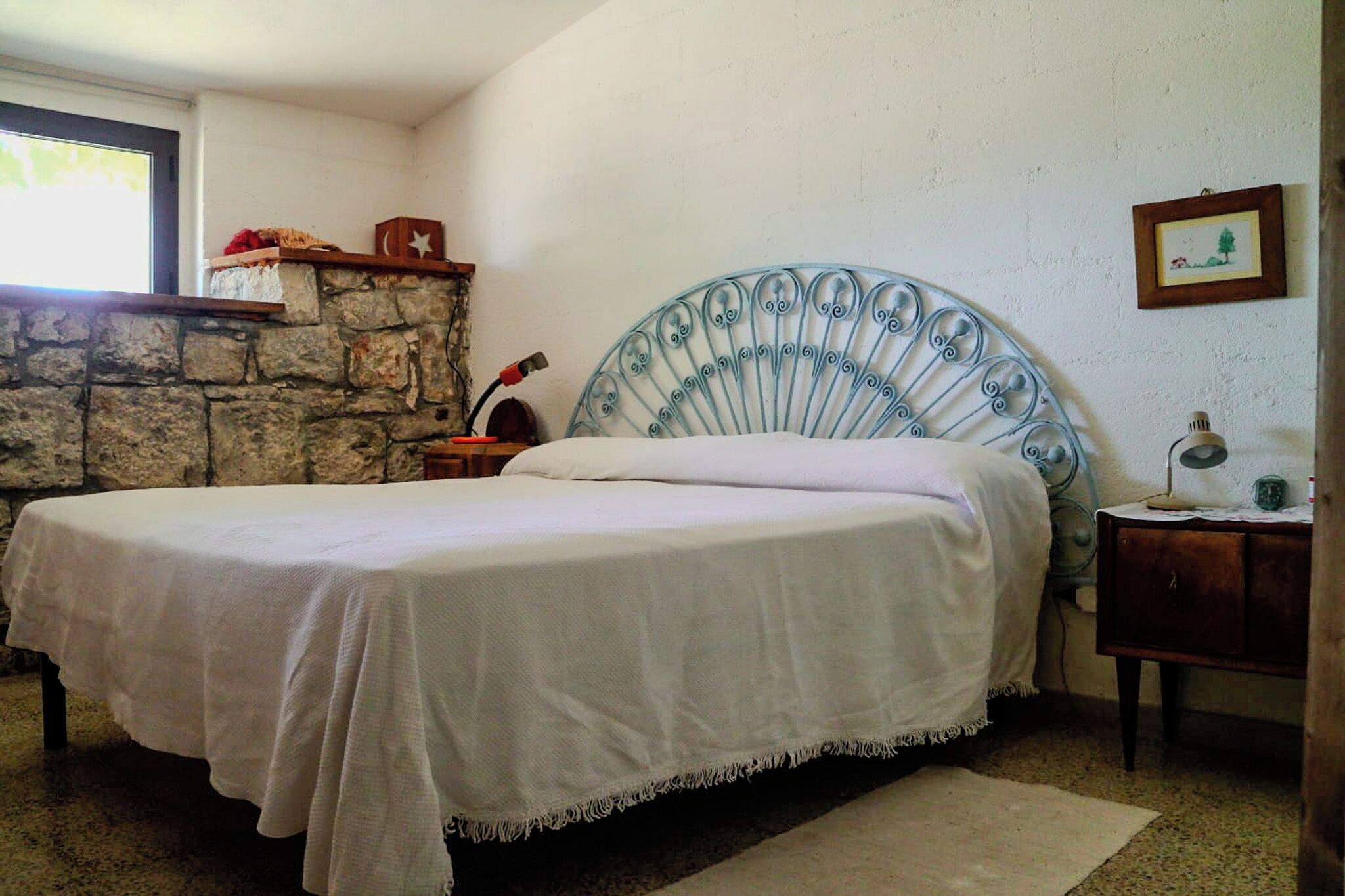 Uma cama com cabeceira de metal, parede de pedra, uma pequena mesa com abajur e um quadro na parede.