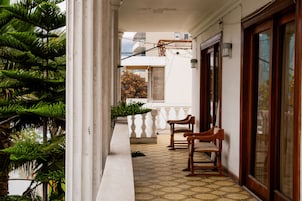 Uma varanda com piso de azulejos, cadeiras de madeira, um vaso com planta e vista para um edifício ao fundo.