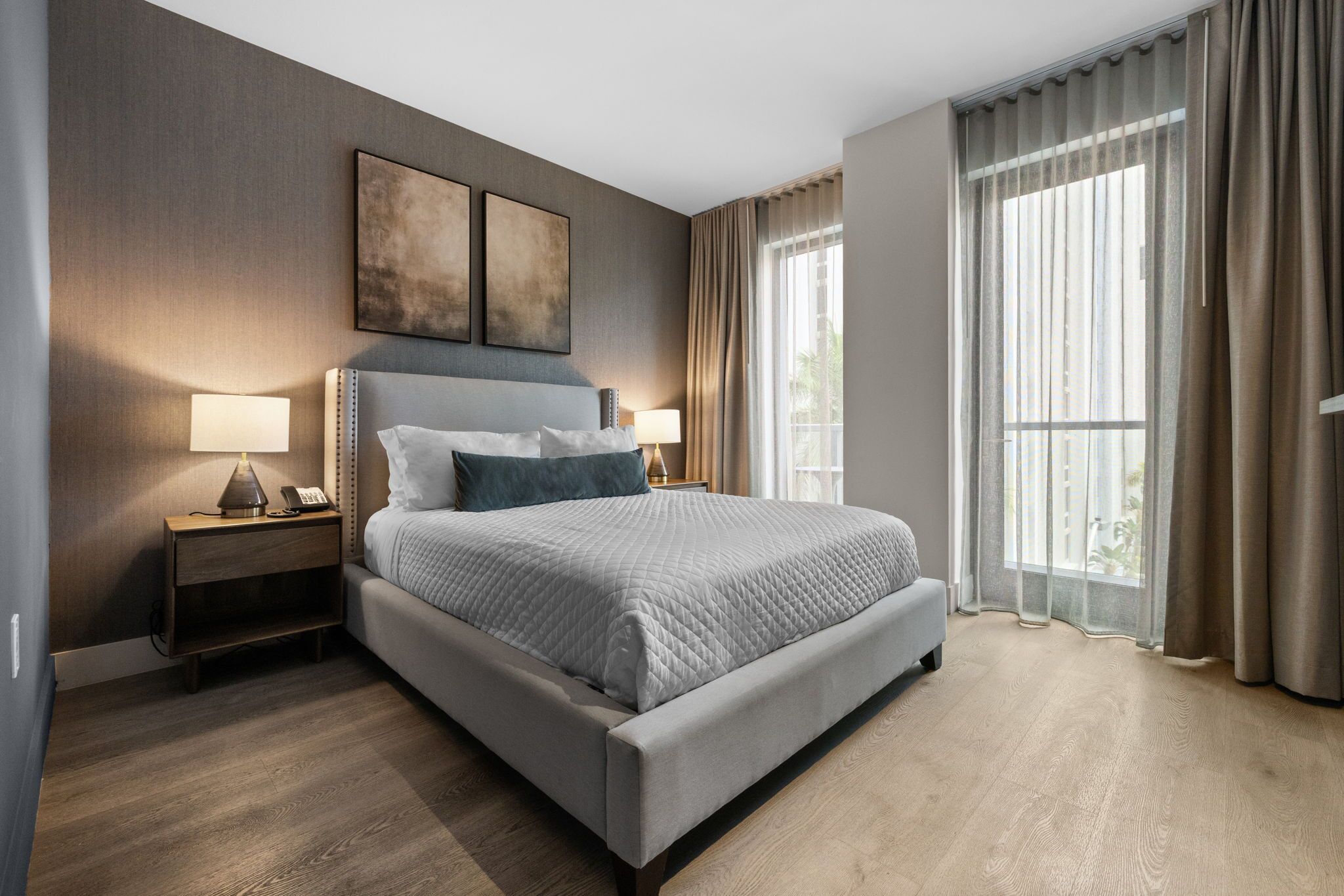 Quarto de hotel moderno com uma cama grande, duas mesas de cabeceira com luminárias, cabeceira com arte e uma janela ampla com cortinas.