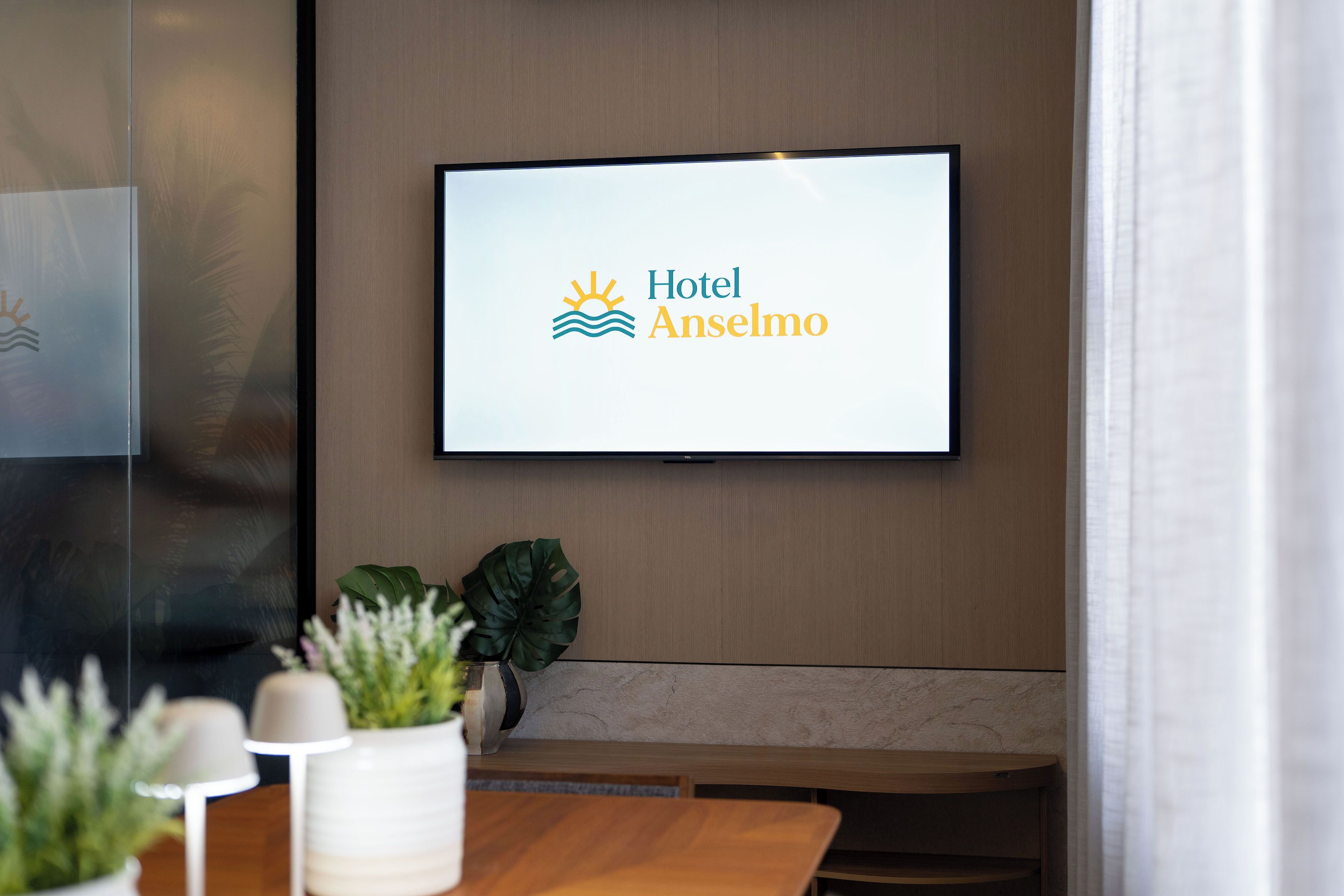 Uma área de recepção de hotel com uma grande tela exibindo o logotipo do Hotel Anselmo, um balcão de madeira e uma planta em vaso.