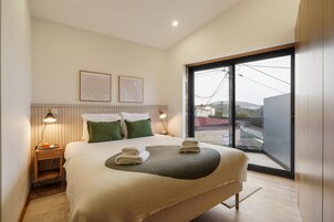 Quarto moderno com uma cama grande, duas luminárias de cabeceira, vista através de uma janela ampla e duas imagens emolduradas na parede.