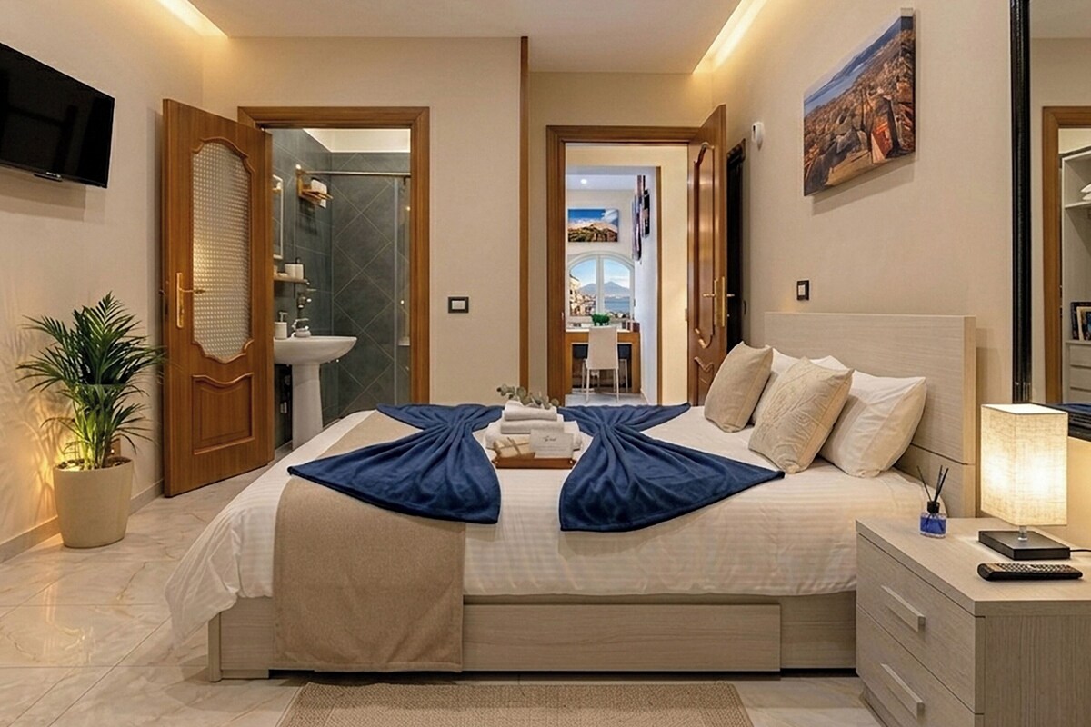 Quarto de hotel com cama, mesa de cabeceira, luminária, TV e vista para a cidade pela janela.