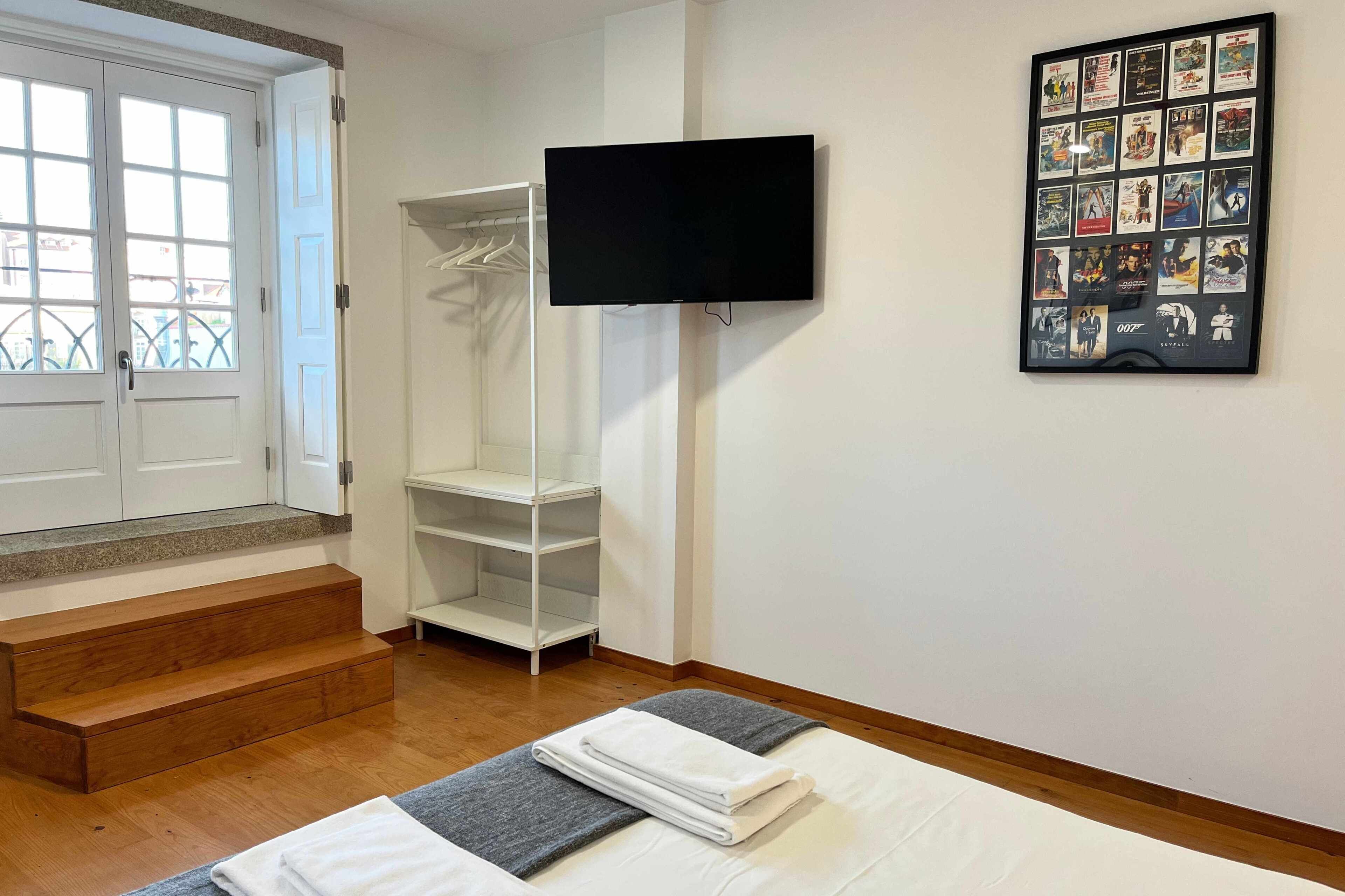 Quarto de hotel com uma cama, televisão fixada na parede, guarda-roupa e escada que dá para a porta.