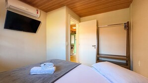 Uma cama de solteiro com colchão cinza e roupa de cama branca, uma TV de tela plana fixada na parede e teto de madeira.