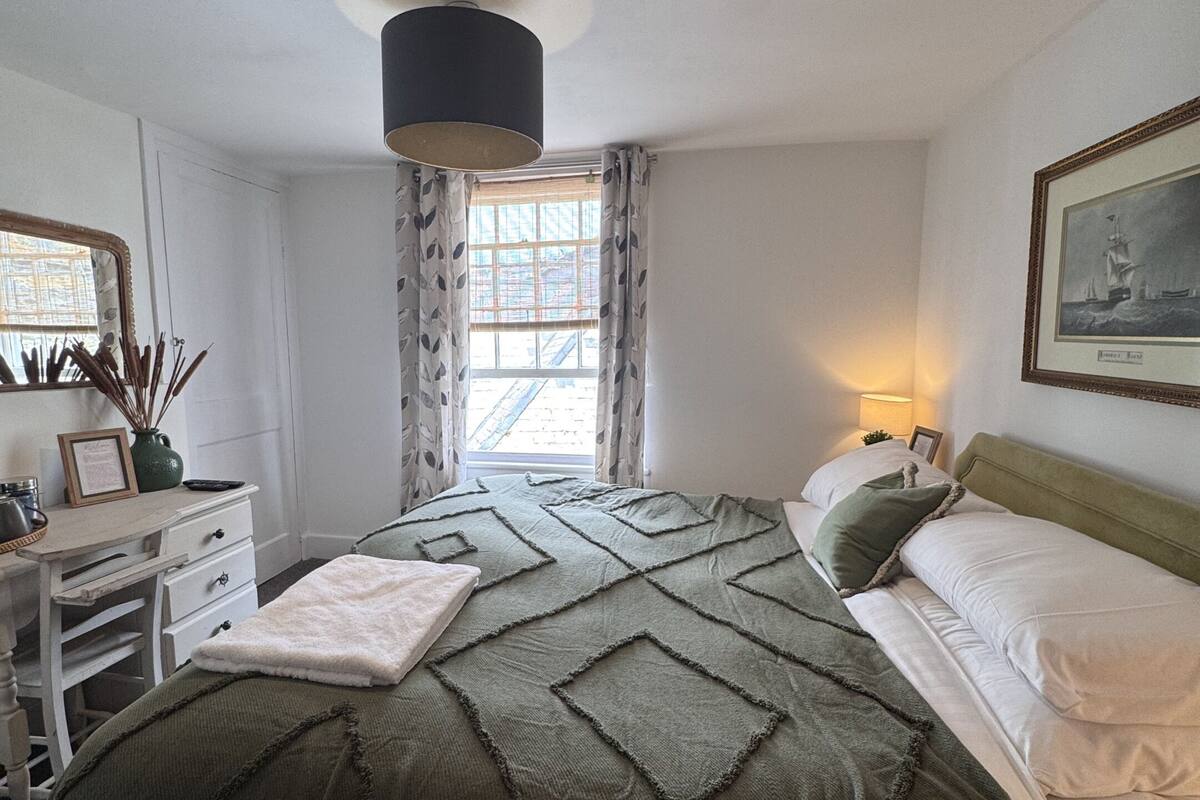 Um quarto com cama, escrivaninha, abajur, janela com cortinas e um quadro na parede.
