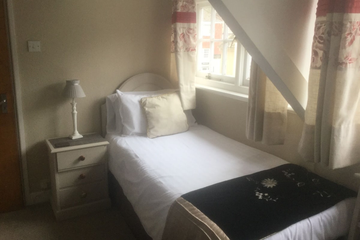 Quarto com uma cama de solteiro, mesa de cabeceira com abajur, janela com cortinas e vista para prédios.
