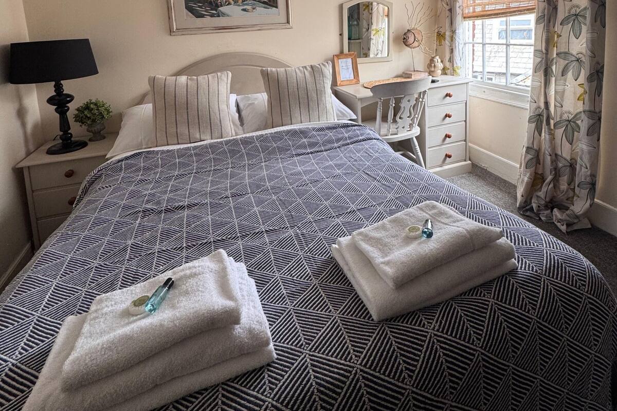 Quarto com cama, mesa de cabeceira, abajur, cômoda, espelho e janela com cortinas.
