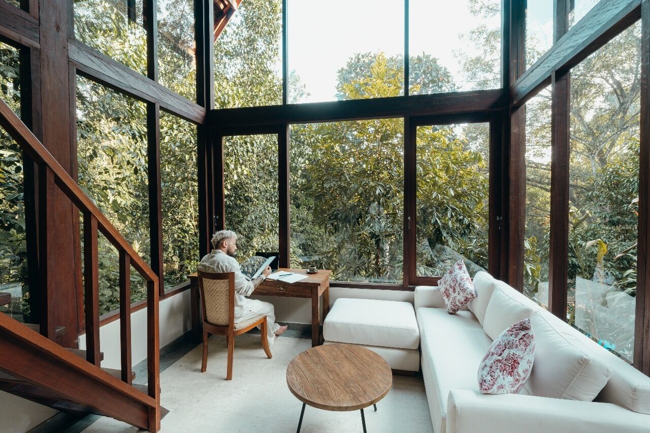 Um quarto espaçoso com um amplo painel de vidro, escada de madeira, sofá branco, mesa de madeira redonda e uma pessoa usando um laptop.