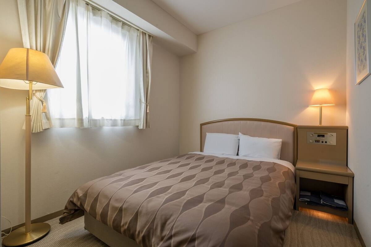 Quarto de hotel com cama, mesa de cabeceira com abajur, janela com cortinas e painel de controle fixado na parede.