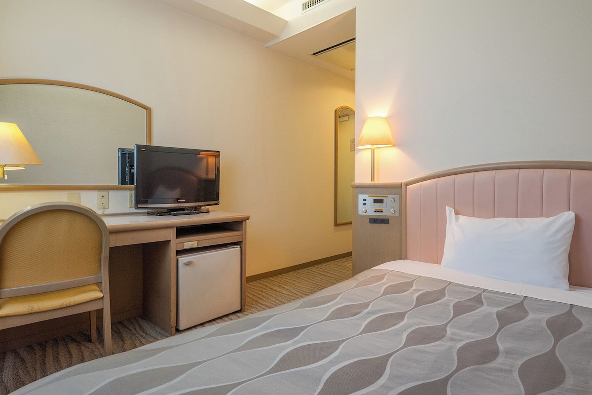 Quarto de hotel com cama, escrivaninha com televisão, cadeira, pequeno frigorífico e mesa de cabeceira com abajur.