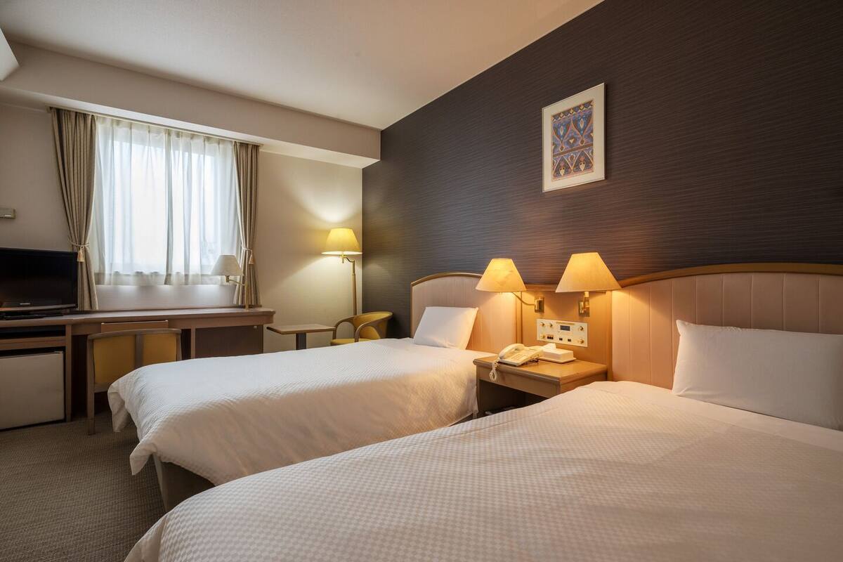 Quarto de hotel com duas camas, uma escrivaninha, televisão e uma luminária.