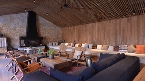 Uma sala de estar ampla, com parede de pedra, teto de madeira e lareira embutida. Há diversas opções de assentos, incluindo sofás, cadeiras e uma mesa de centro.