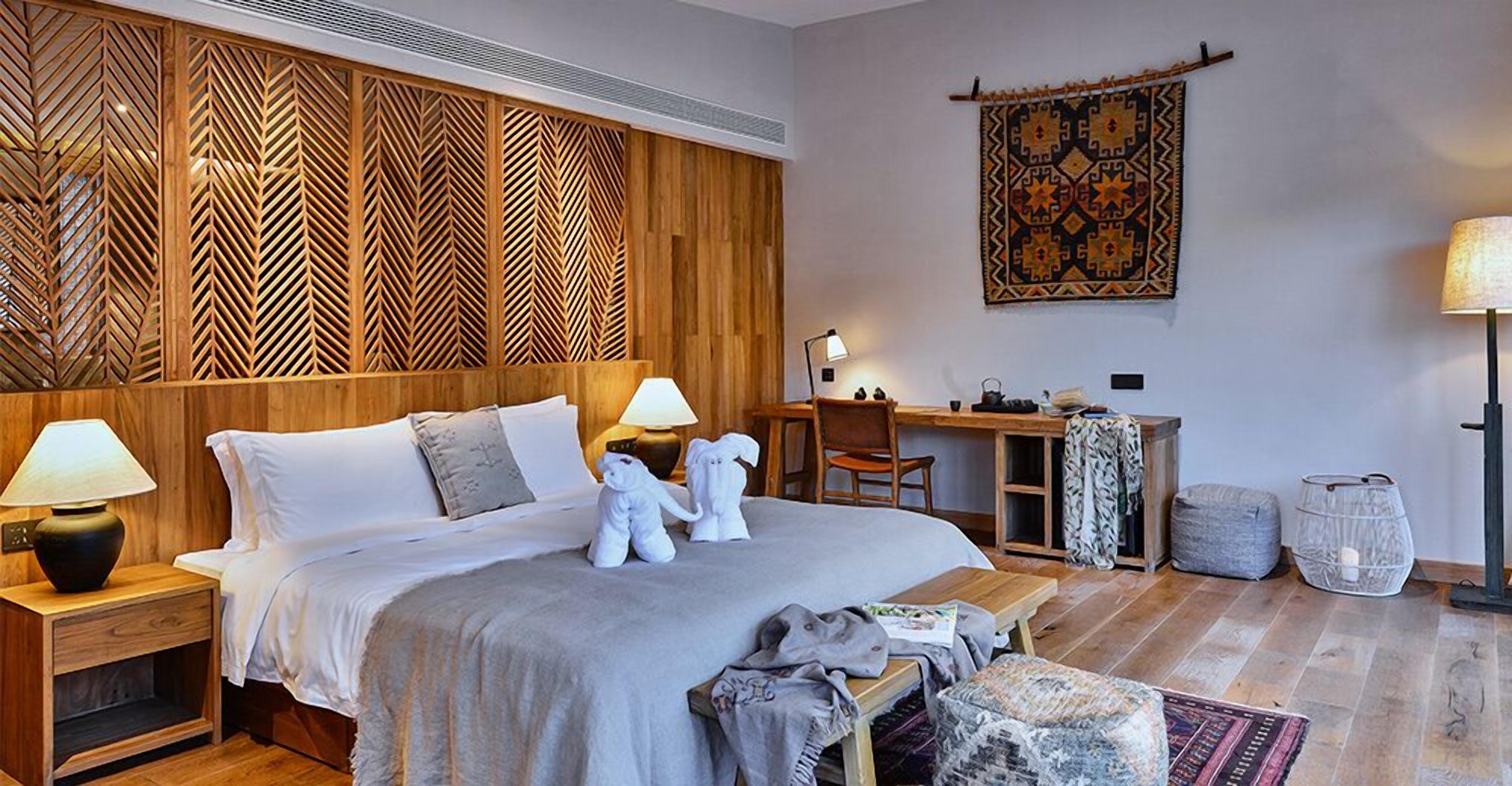 Quarto de hotel com cabeceira de madeira, escrivaninha, cadeira, cama com roupa de cama branca e um enfeite de parede.
