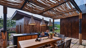 Uma área de cozinha externa com mesa de madeira, luminárias pendentes e um painel de fundo rústico em madeira.