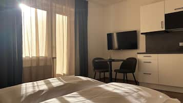 Quarto de hotel com cama, escrivaninha, duas cadeiras, televisão e cozinha compacta.