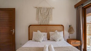 Cama bem arrumada com edredom branco e duas toalhas enroladas, um painel de macramé na parede e um criado-mudo de vime com uma luminária.