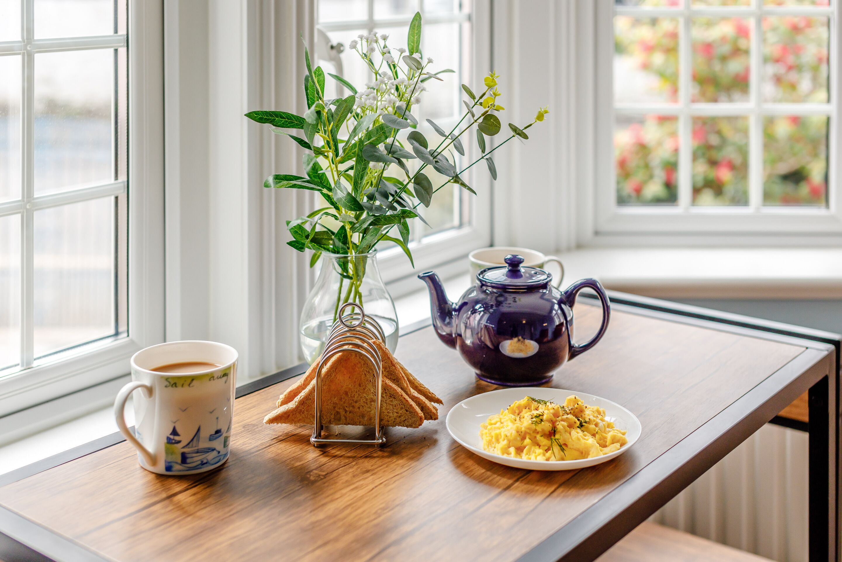 Uma mesa com uma caneca branca, um bule, um prato de ovos mexidos e um vaso de vidro com plantas verdes.