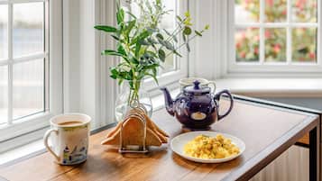 Uma mesa com uma caneca branca, um bule, um prato de ovos mexidos e um vaso de vidro com plantas verdes.