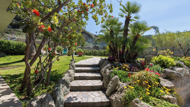 Uma escadaria de pedra leva a um edifício, cercada por vegetação exuberante e plantas floridas.