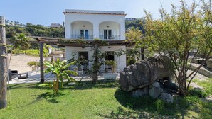 Um edifício branco com duas varandas, rodeado por um jardim com uma grande rocha e uma árvore.