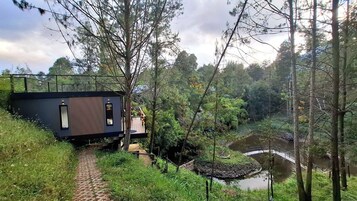 Um edifício moderno em estilo cabana, com andar superior de vidro e andar inferior de madeira, localizado numa encosta com um rio e área arborizada ao fundo.