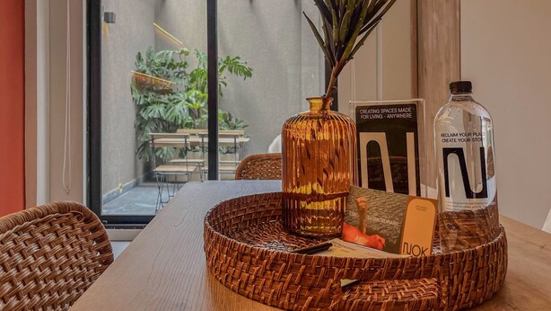 Uma mesa de jantar com uma cesta trançada, um vaso com uma planta, uma garrafa e um cartão.