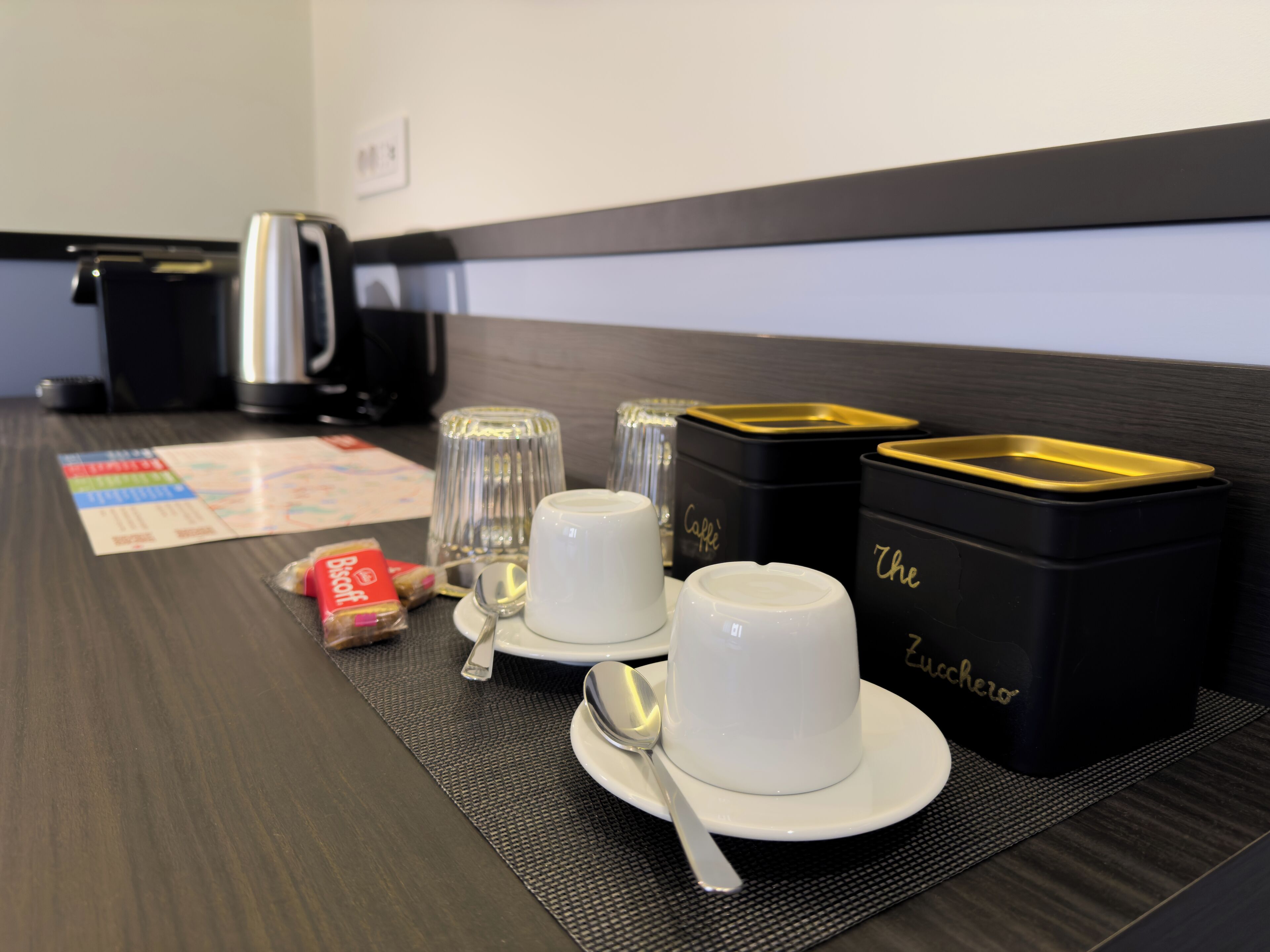 Uma mesa com uma cafeteira, chávenas, uma colher e uma caixa com a inscrição "The Zucker".