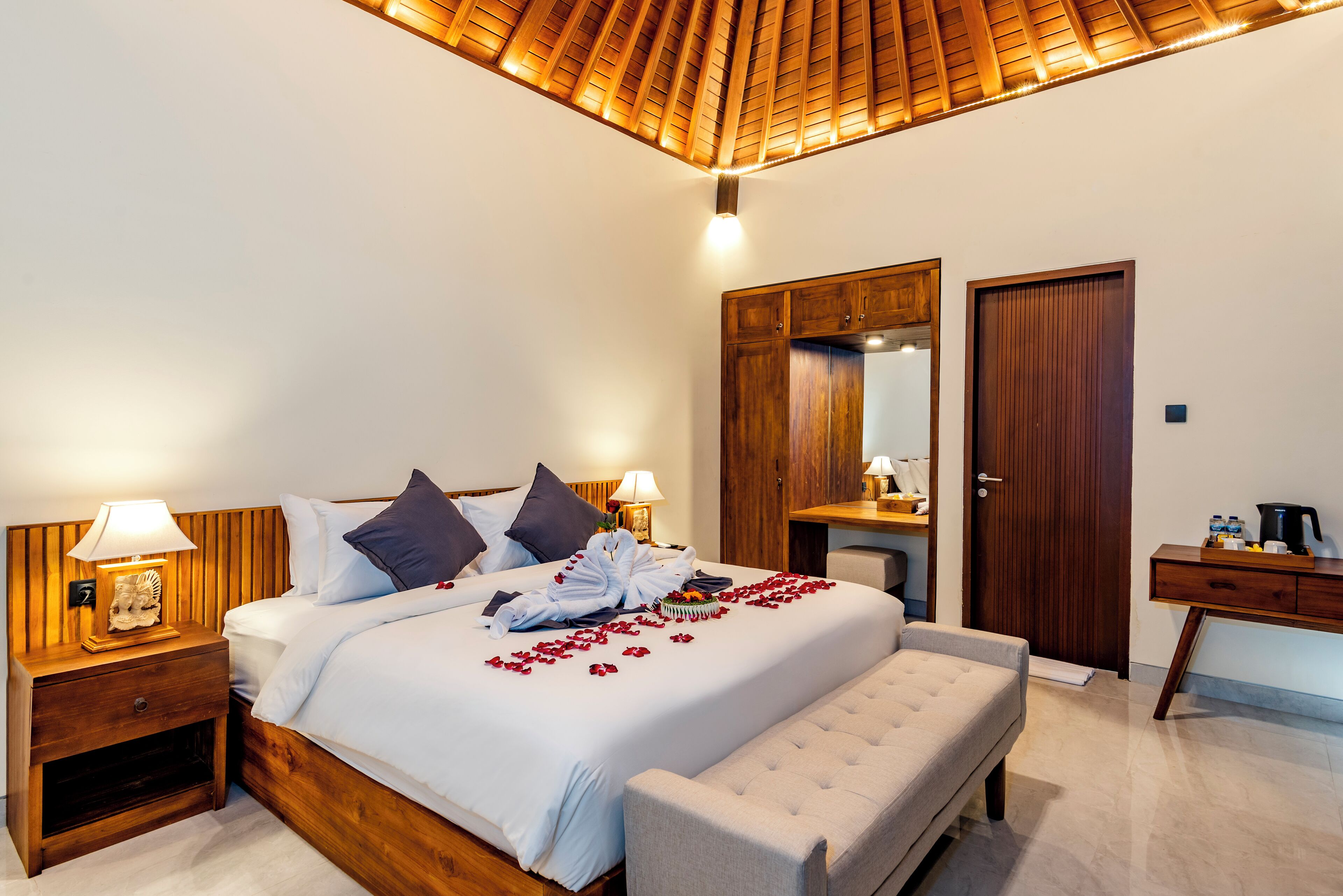 Quarto bem organizado com cabeceira de madeira, mesa de cabeceira com abajur, um banco e guarda-roupa de madeira.