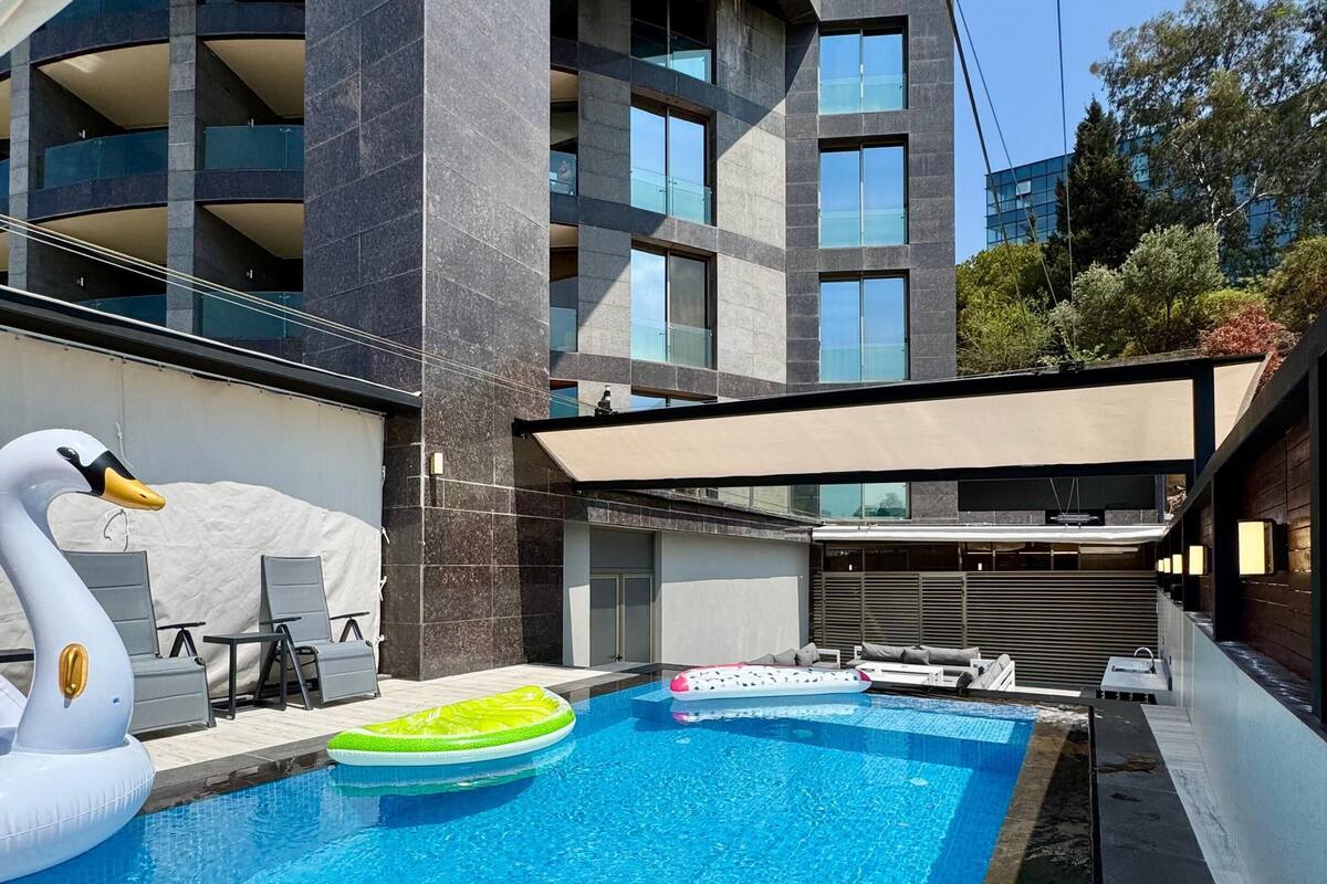 Piscina com cisne inflável e boia verde, cercada por arquitetura moderna.