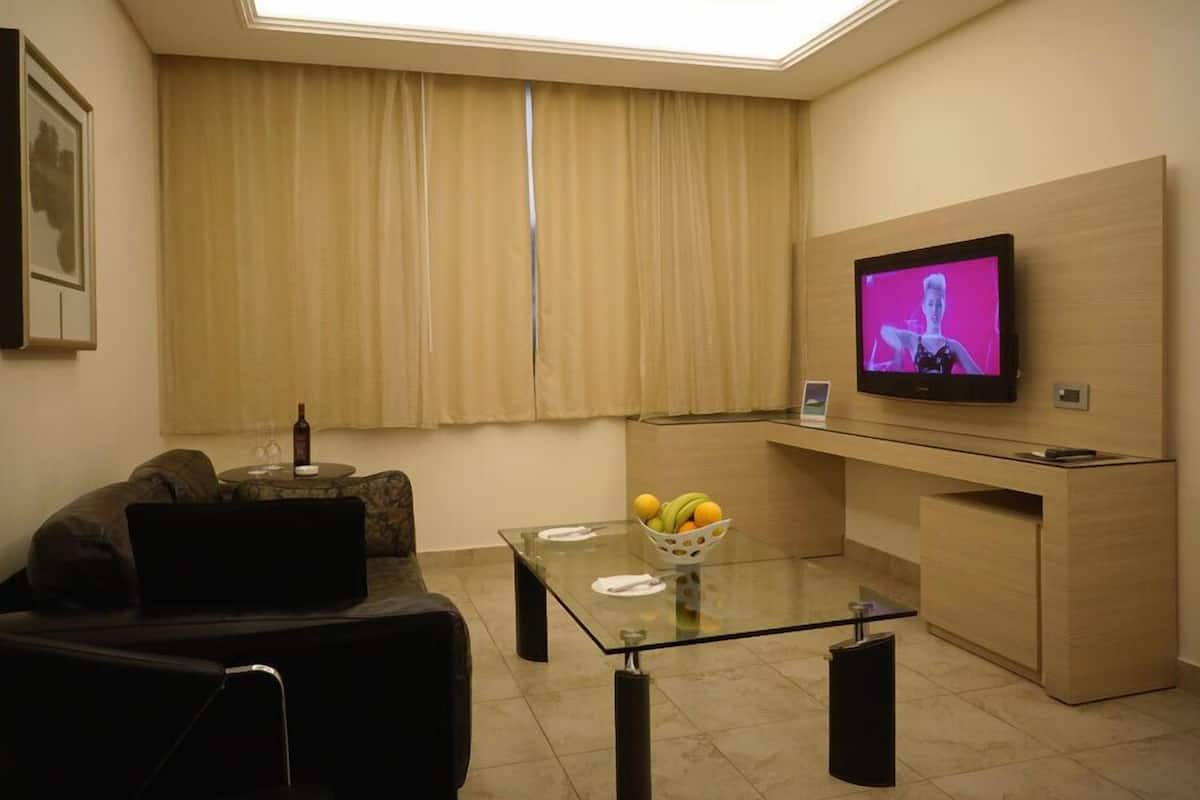 Uma sala de estar moderna com uma mesa de centro de vidro, um sofá, uma TV fixada na parede e uma janela com cortinas.
