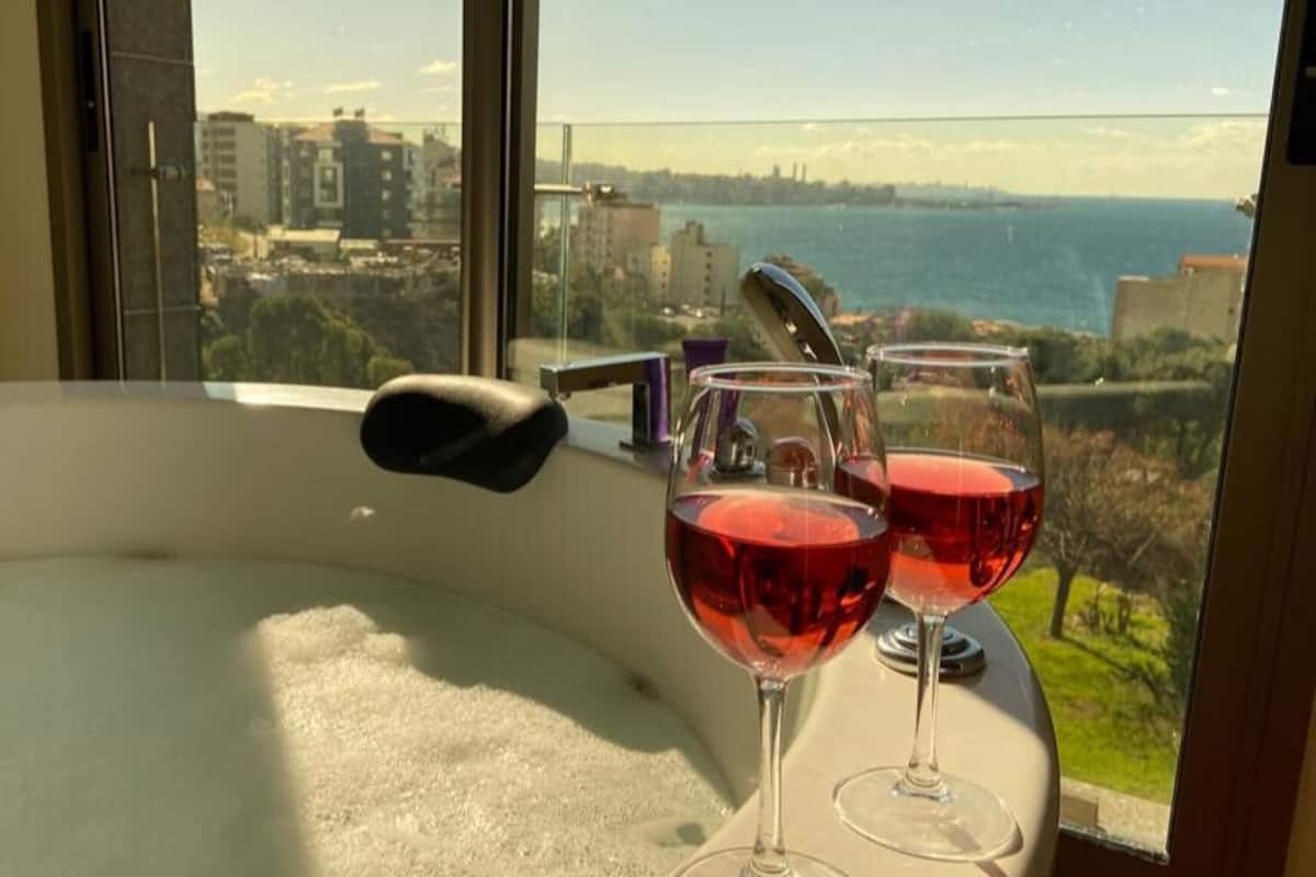 Banheira de hidromassagem com dois copos de vinho tinto e vista para a paisagem urbana e o mar.