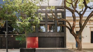 Um edifício moderno com uma porta vermelha, uma árvore em frente e uma rua no primeiro plano.