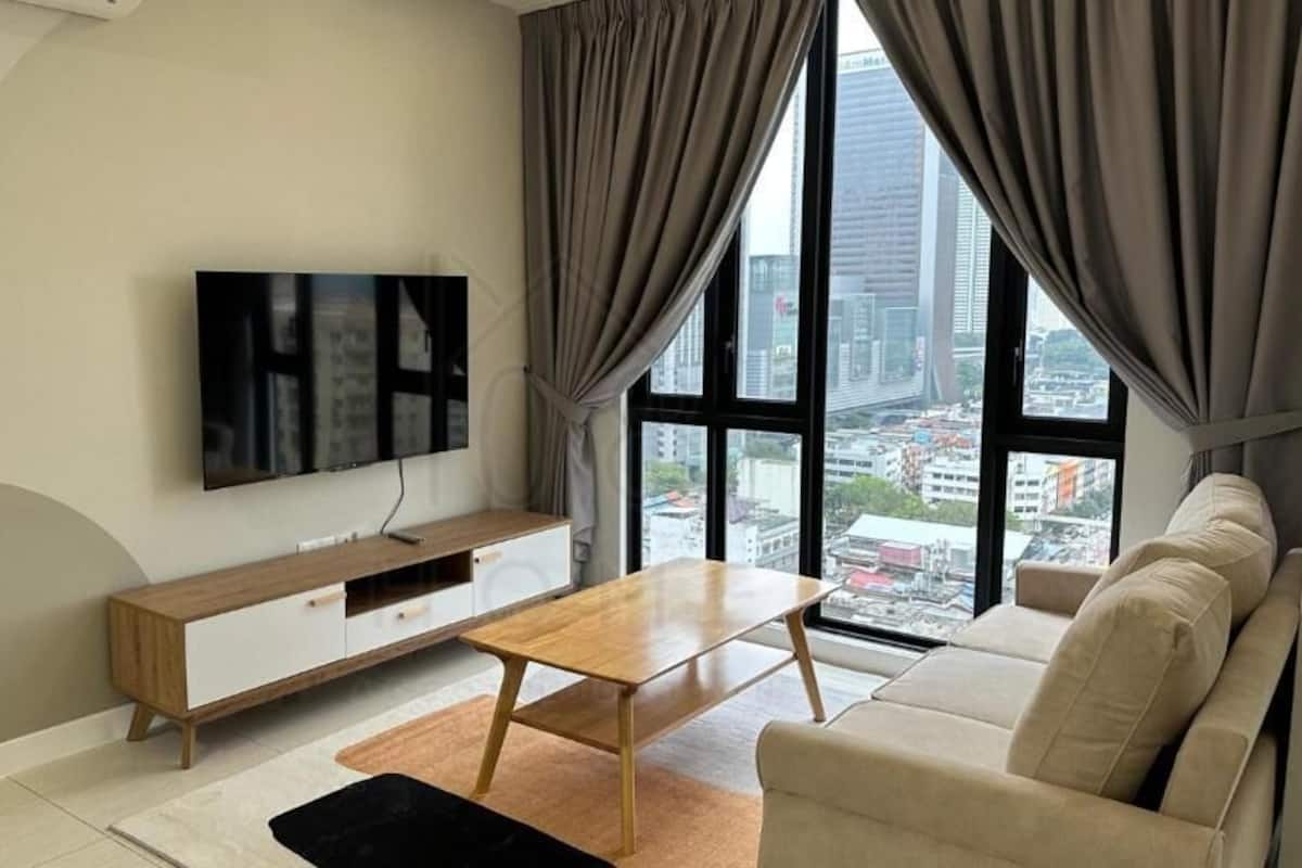 Uma sala de estar moderna com televisão na parede, mesa de centro de madeira, sofá bege e uma janela grande com vista para a cidade.