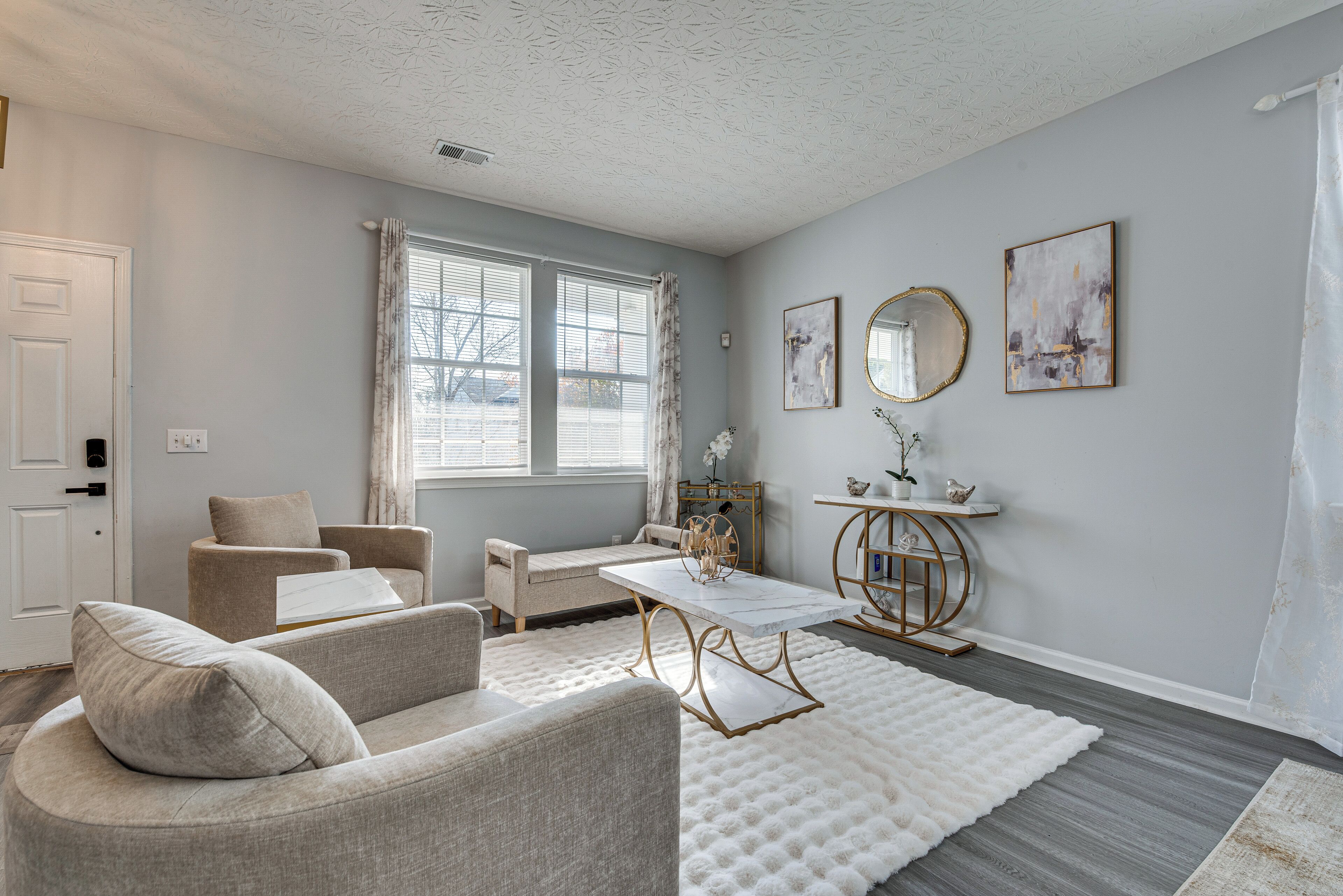 Uma sala de estar moderna com um sofá, poltrona e mesa de centro. Há uma janela com cortinas, um espelho e uma pequena mesinha lateral com objetos decorativos.