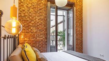 Quarto com parede de tijolos, cama com almofadas amarelas e bege, luminária pendente e vista através de uma porta de vidro.