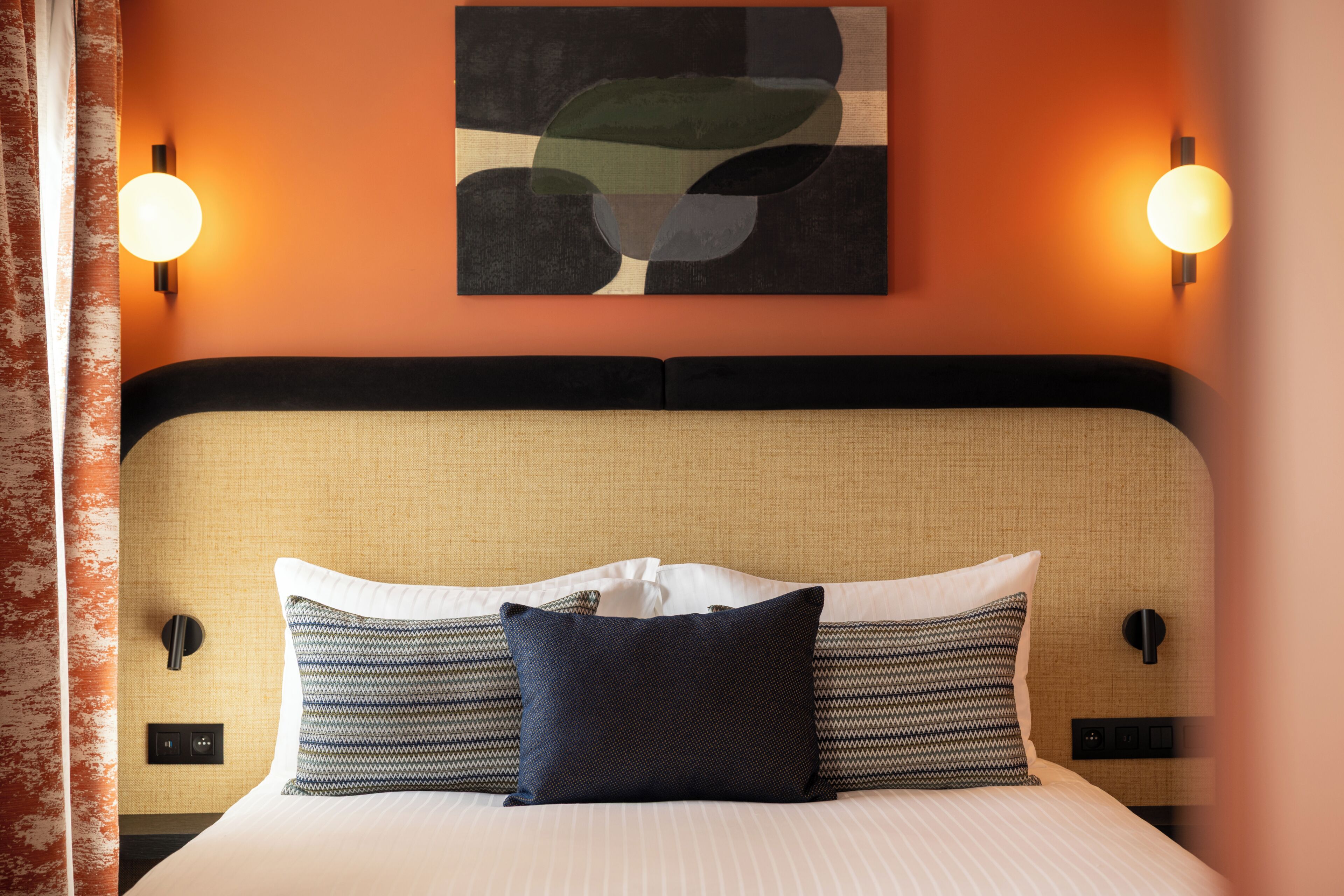 Quarto de hotel com cama, duas arandelas, cabeceira e uma obra de arte na parede.