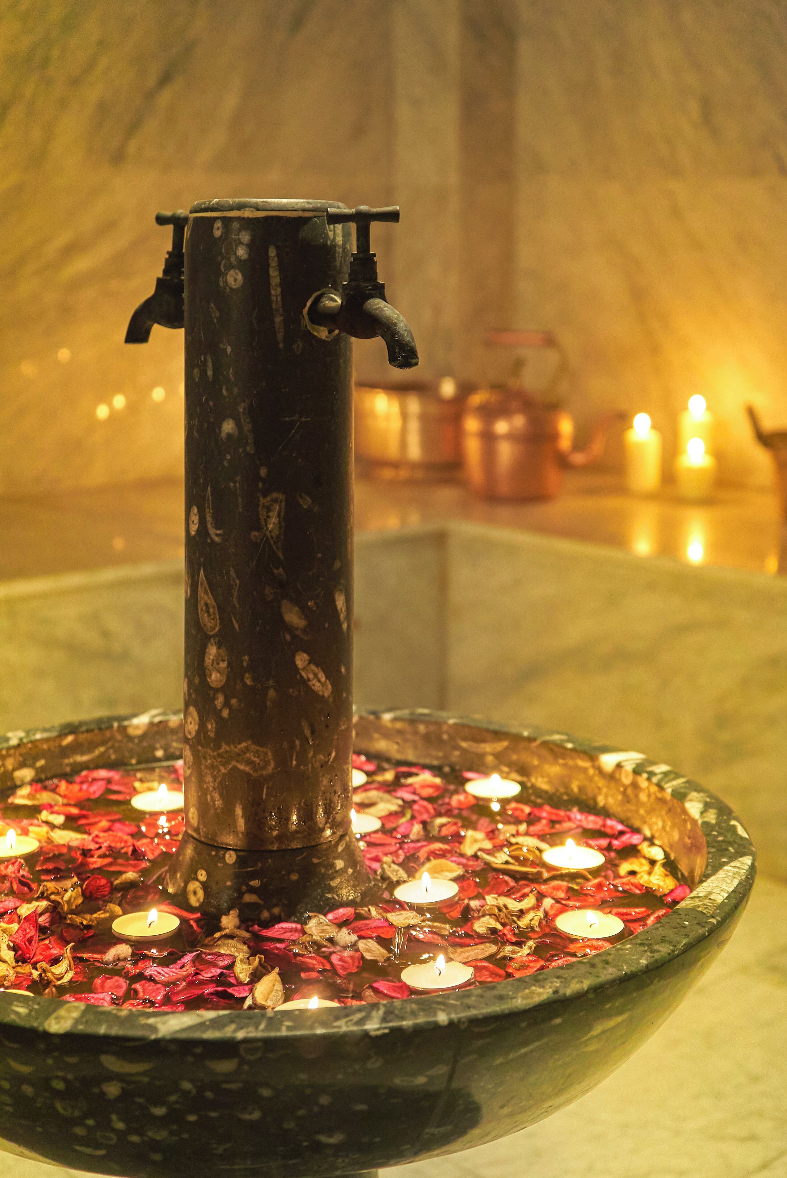 Uma bacia de pedra com uma coluna central, rodeada por velas e pétalas de rosa.
