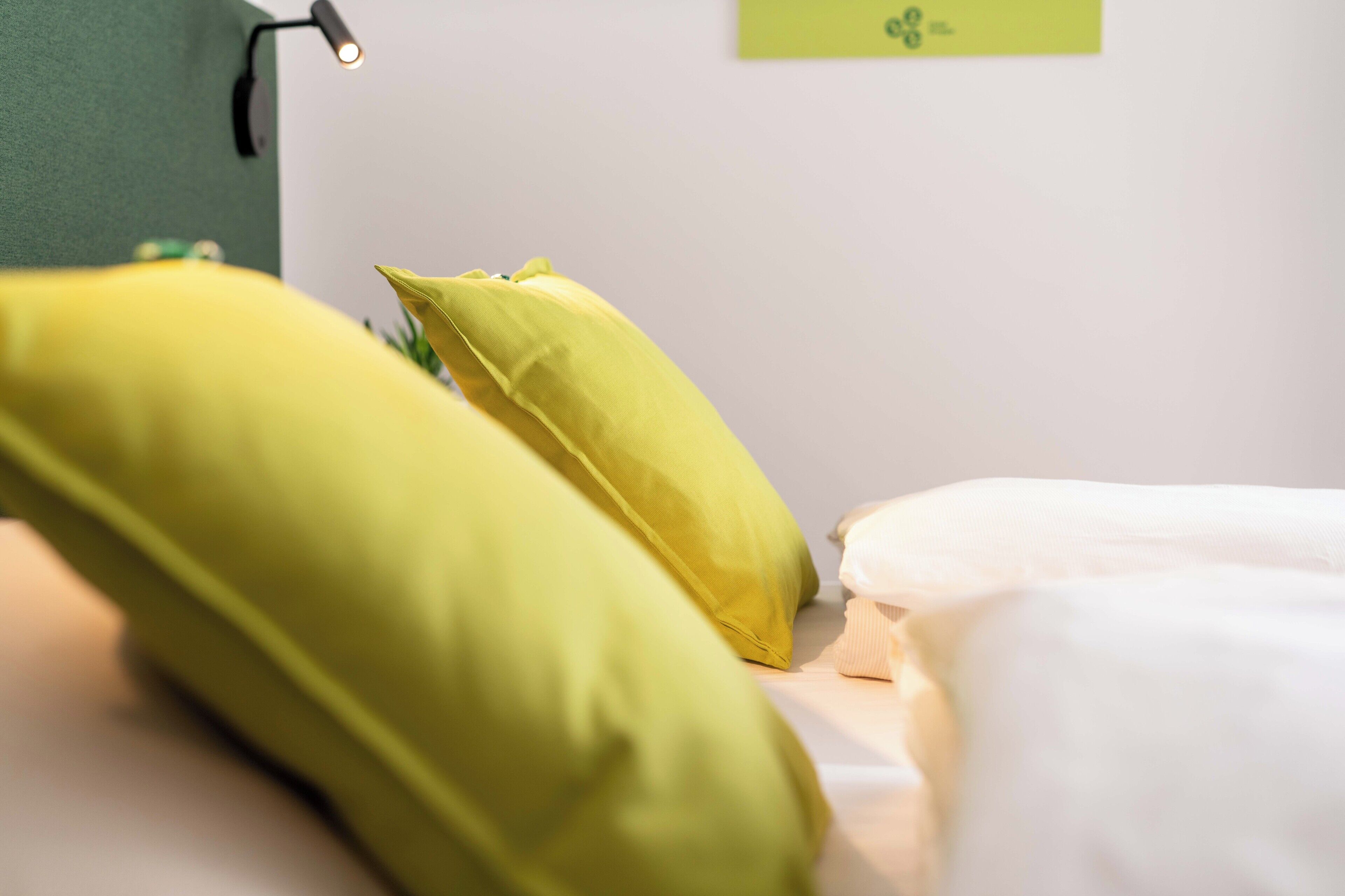 Uma cama com almofadas verdes e brancas, um abajur e uma parede verde com uma pequena placa verde.