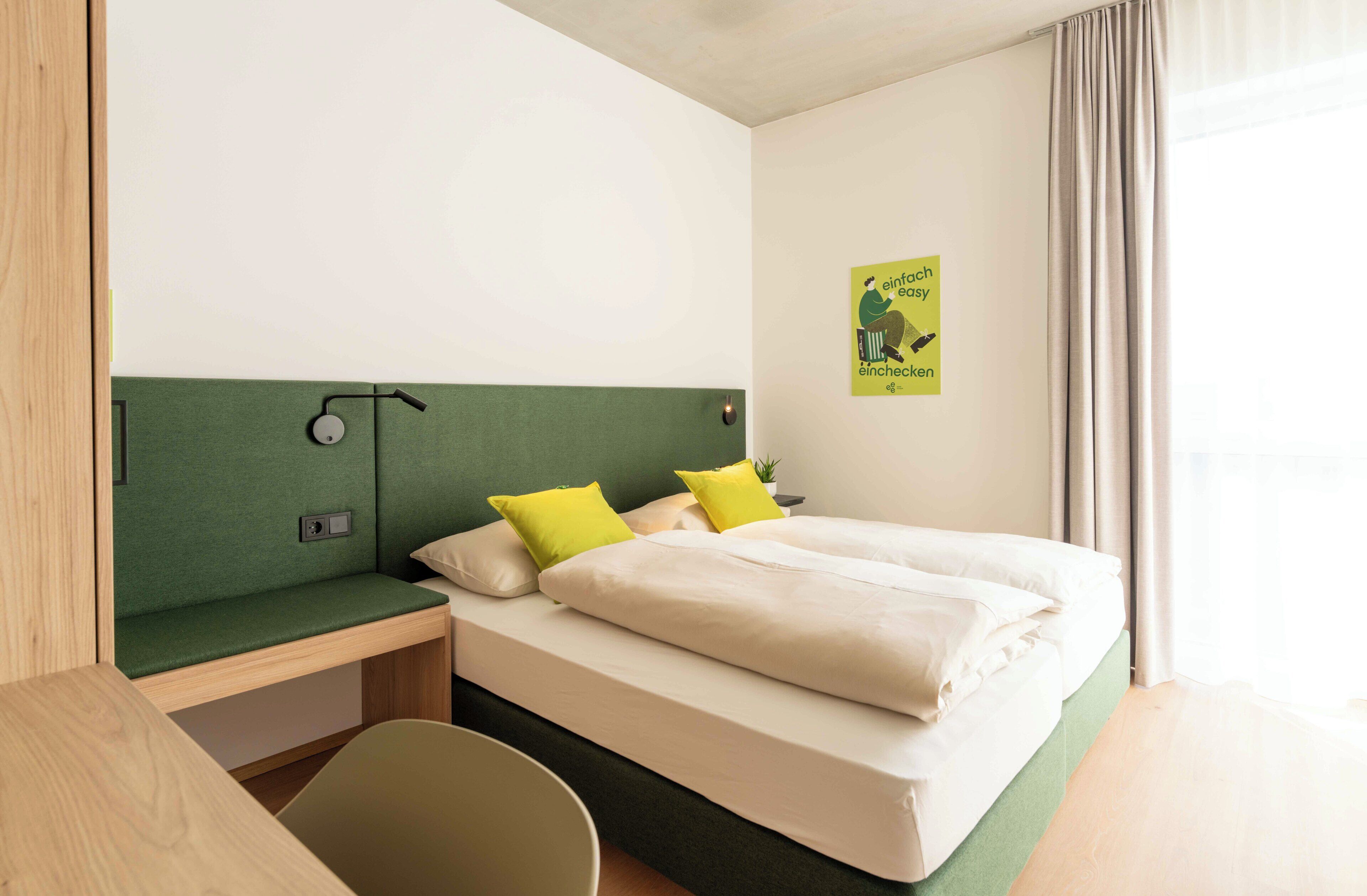 Quarto de hotel moderno com um banco estofado verde, cama com roupa de cama branca e almofadas amarelas, uma pequena mesa de cabeceira e um cartaz na parede.