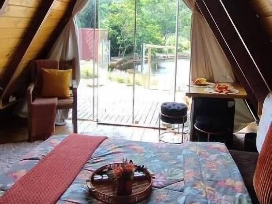 Um quarto de sótão acolhedor com teto inclinado, uma cama com colcha florida, uma mesinha com cesto e vista para um jardim através de uma porta de vidro.