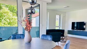 Sala de estar moderna com uma mesa redonda, cadeiras azuis, televisão de tela plana e uma janela grande com vista para o verde.