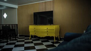 Quarto com piso quadriculado, cômoda amarela e televisão de tela plana fixada em uma parede de madeira.